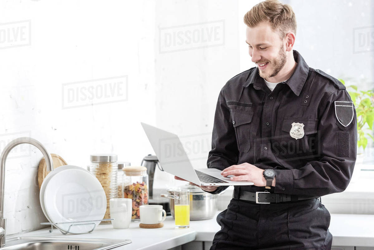 smiling police officer standing and using laptop at kitchen - Royalty ...