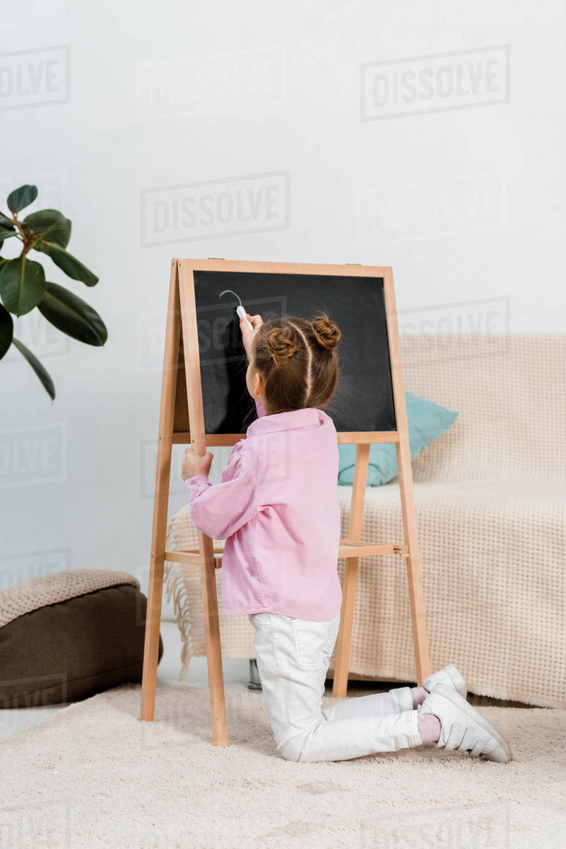 Back view of cute kid kneeling on carpet and writing on chalkboard ...