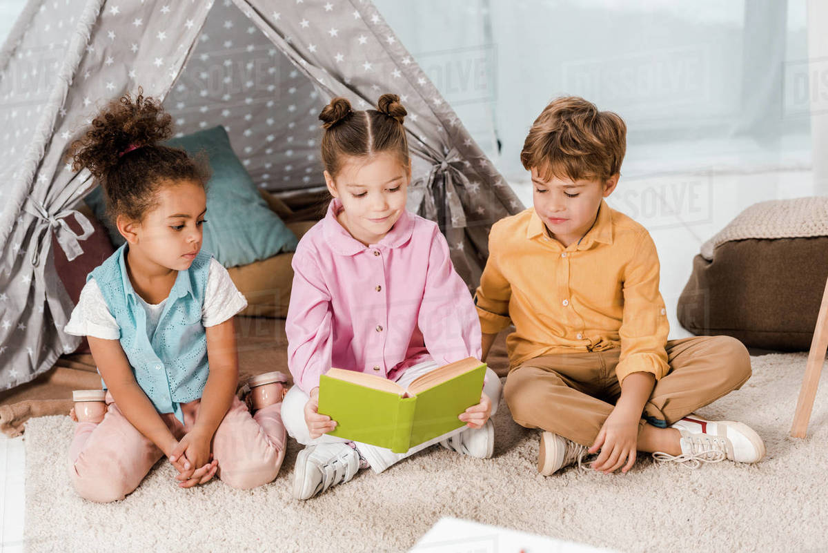 Adorable little children sitting on carpet and reading book together ...