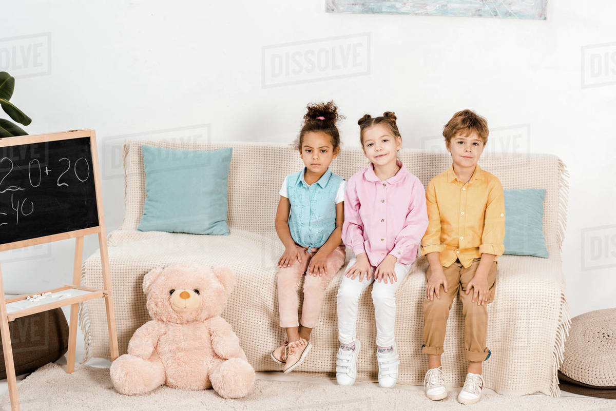 Adorable little multiethnic kids sitting on couch and looking at camera ...