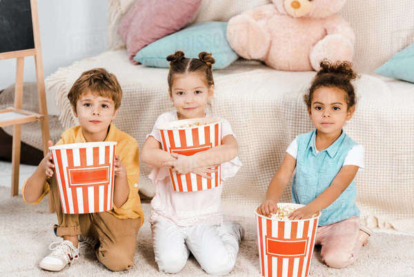 Adorable multiethnic kids holding boxes with popcorn and looking at ...