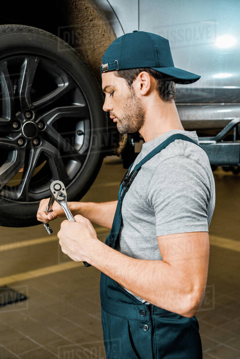 Side view of male auto mechanic in working overall standing with wheel ...