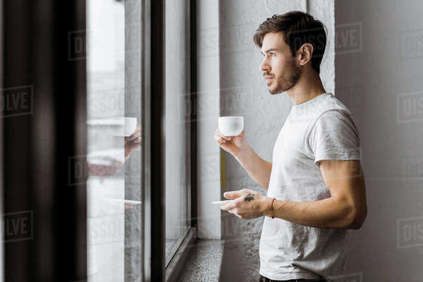 side view of young man holding cup of coffee and looking at window in ...