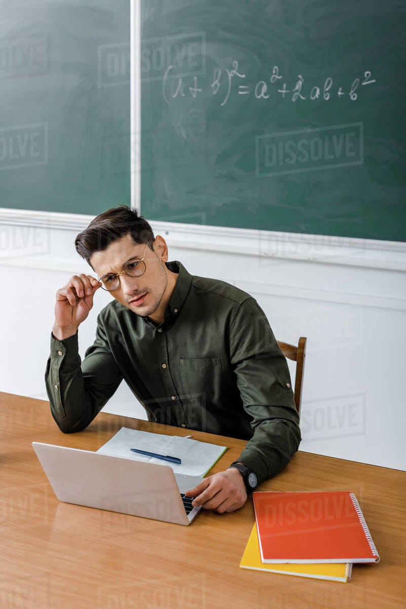 Pensive male teacher in glasses sitting and using laptop at desk in ...