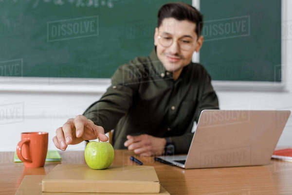 Male teacher in glasses sitting at computer desk and reaching for apple ...