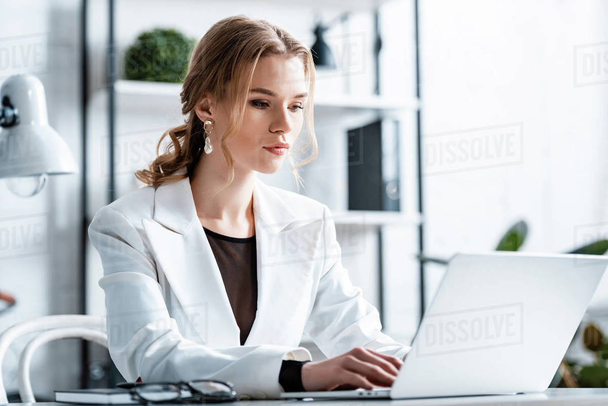 Serious businesswoman in formal wear sitting at desk and typing on ...