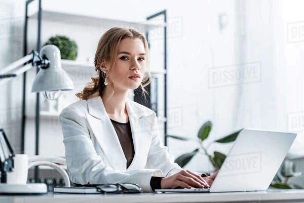 Serious businesswoman in formal wear sitting at desk, looking at camera ...