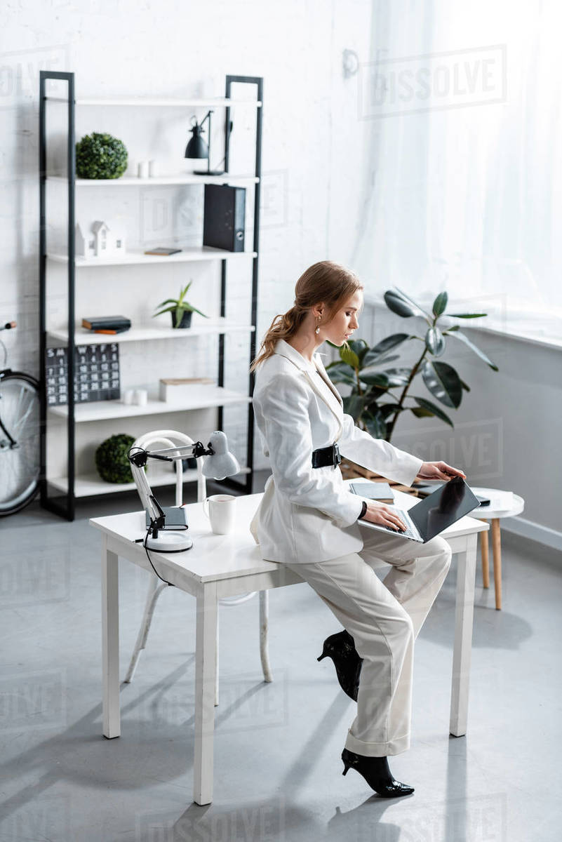 Focused businesswoman in white formal wear sitting on desk and using ...