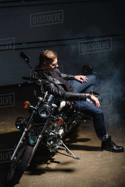 Side view of handsome guy in sunglasses sitting on motorcycle in garage ...
