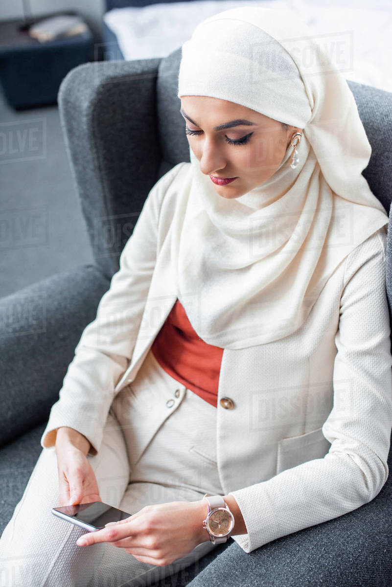Beautiful young muslim woman sitting in armchair and using smartphone ...