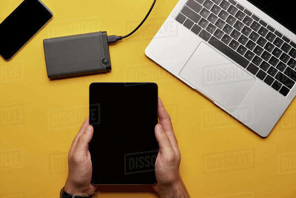 Cropped shot of man using tablet on yellow surface with laptop and ...