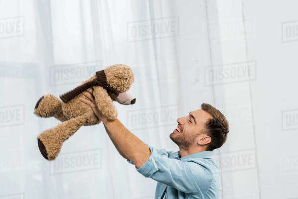 Smiling man holding teddy bear in raising hands in living room ...