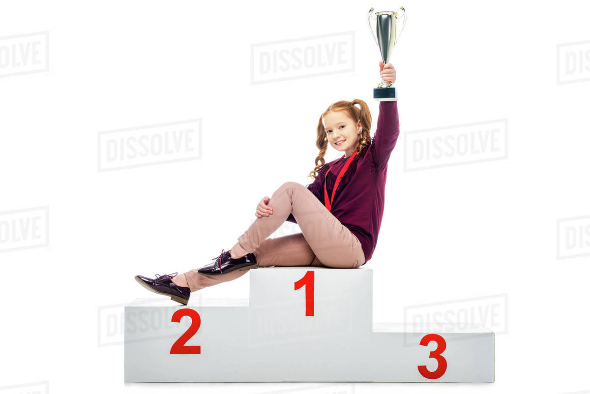 Smiling schoolgirl sitting on winner podium, holding trophy cup in ...