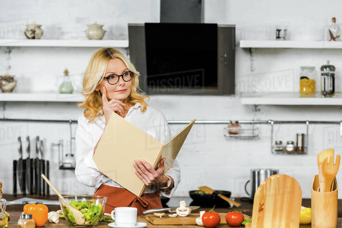 Pensive beautiful mature woman reading recipe book for cooking in ...