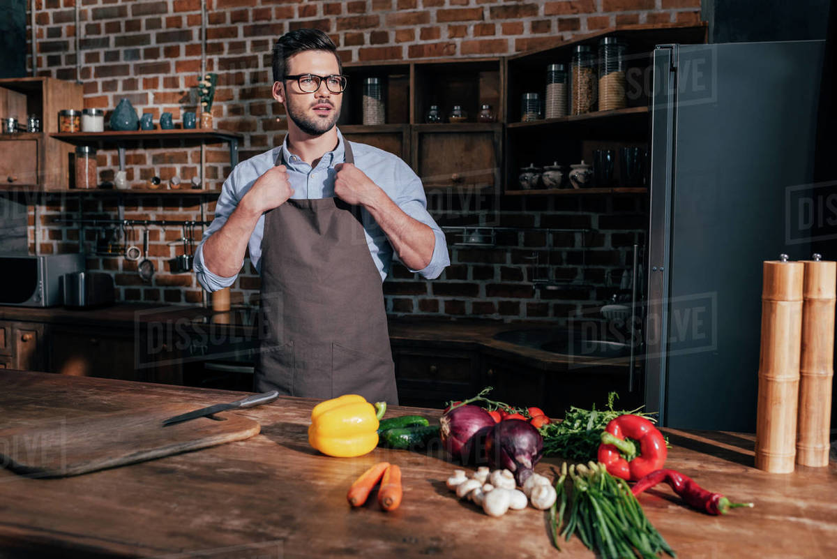 Stylish young man in apron on kitchen with fresh vegetables on table ...