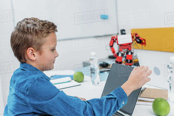 Schoolboy sitting at table with robot model and using laptop during ...
