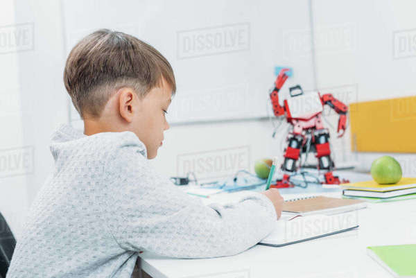 Schoolboy sitting at desk with robot model and writing in notebook ...
