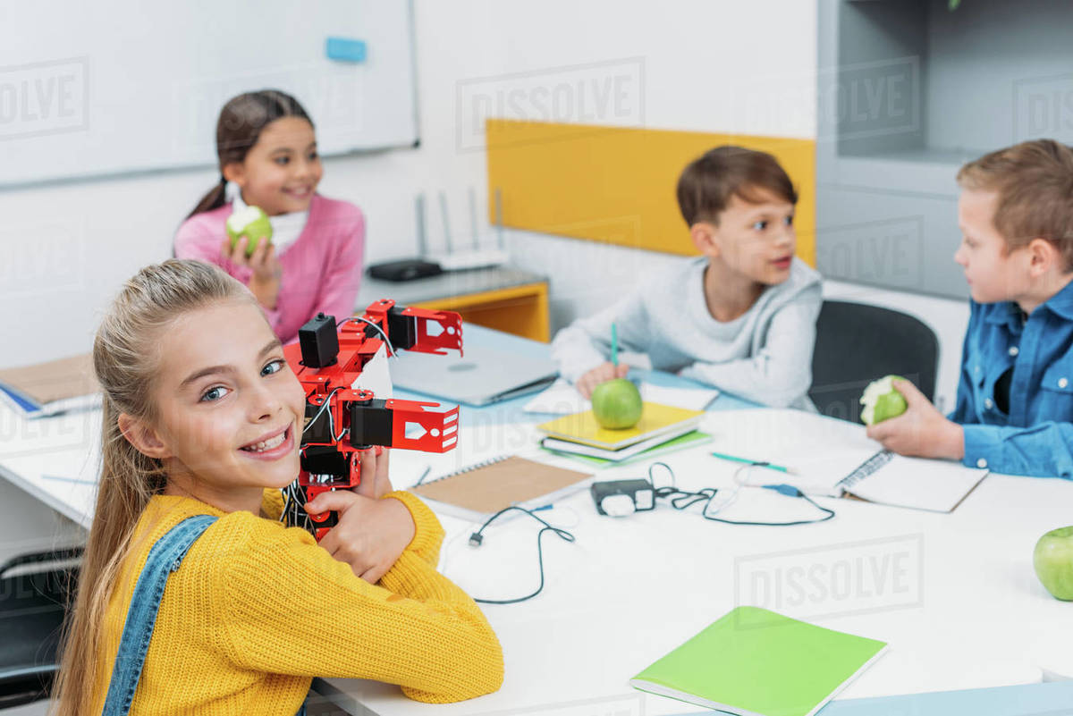 Happy schoolgirl holding robot model while classmates eating apples on ...
