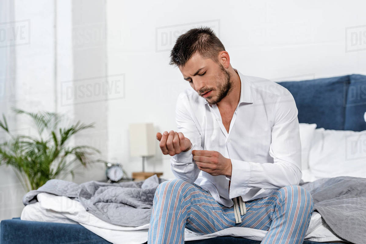 Handsome man sitting on bed and buttoning cuff in weekday morning in ...