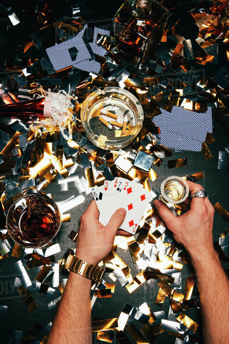 Partial view of man with playing cards and alcohol sitting at table ...