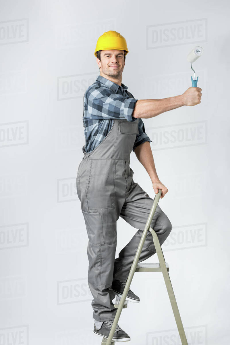 Smiling workman standing on ladder and painting wall with roller ...