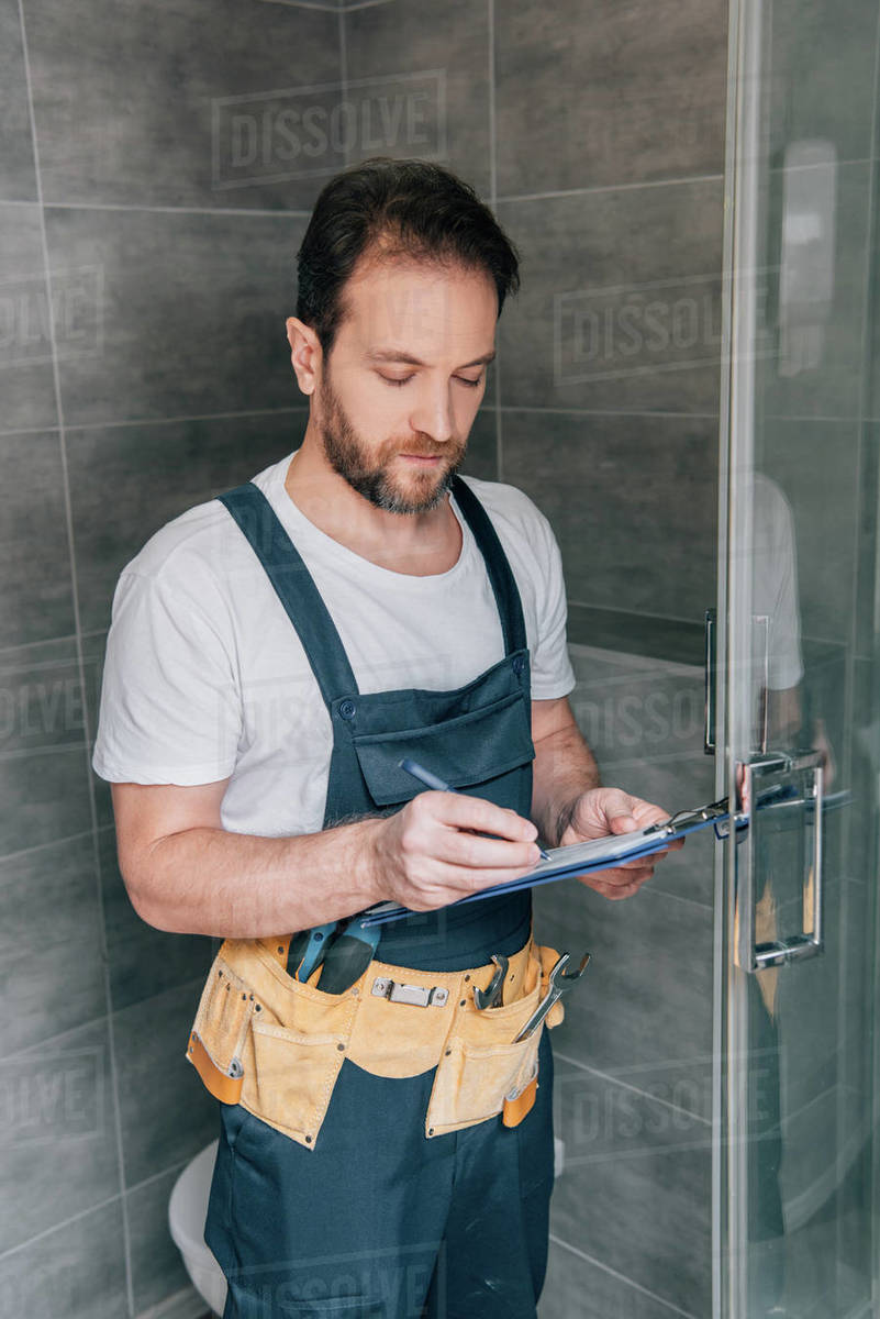 bearded male plumber making notes in clipboard while checking shower in ...