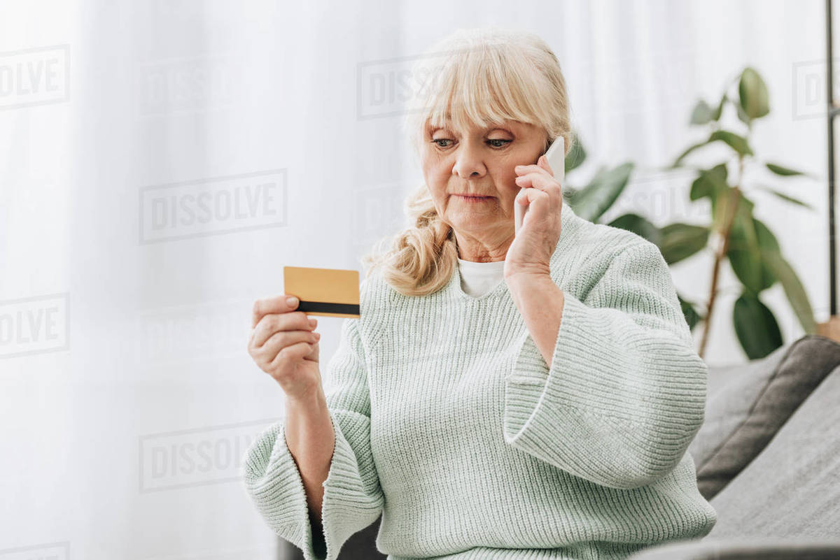 Confused senior woman looking at credit card while talking on ...
