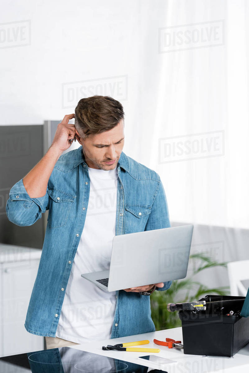 Confused man holding laptop with tool box on table in kitchen - Royalty ...