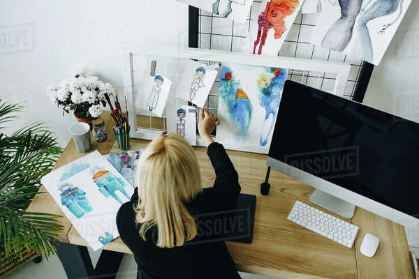 Back view of young blonde fashion designer working with sketches at ...