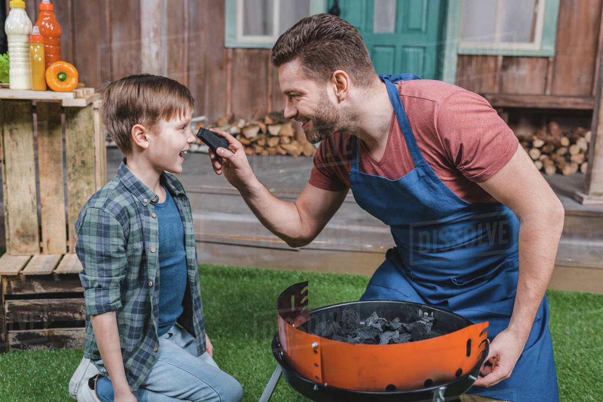 Side view of smiling father and son preparing grill for barbecue ...