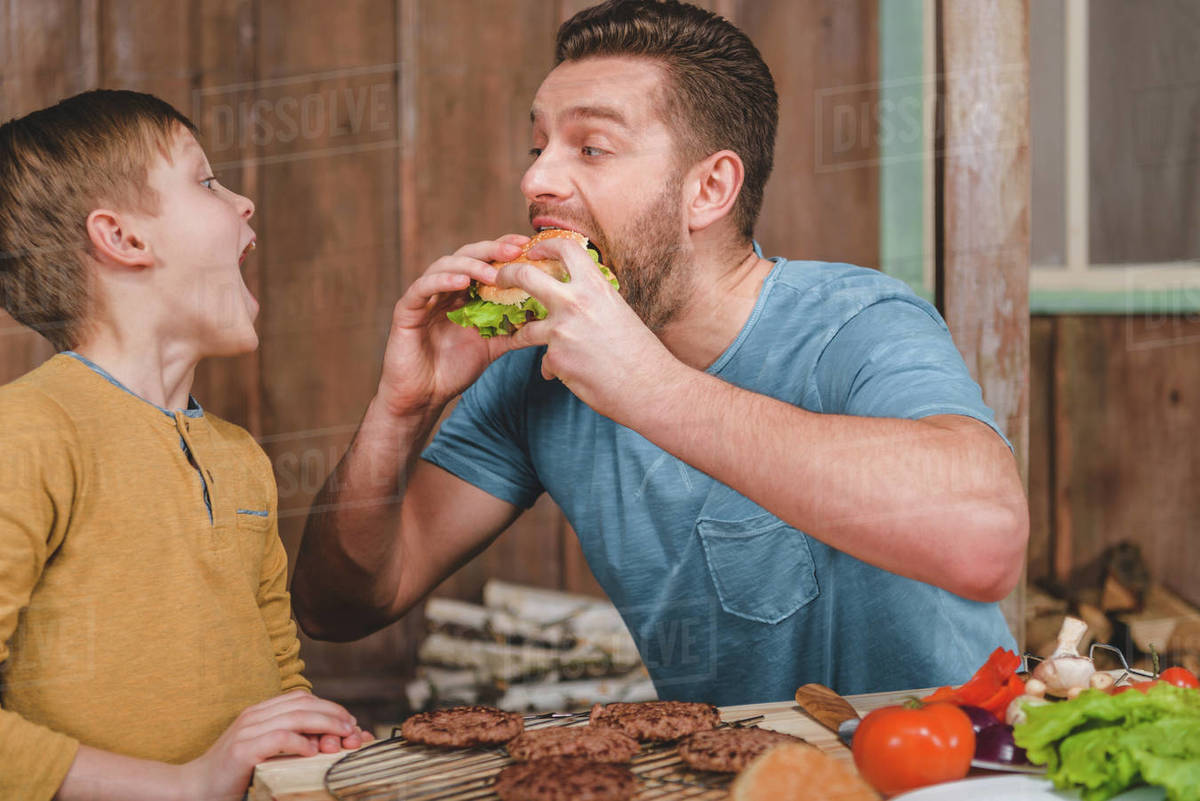 Side view of man eating homemade burger with little son near by ...