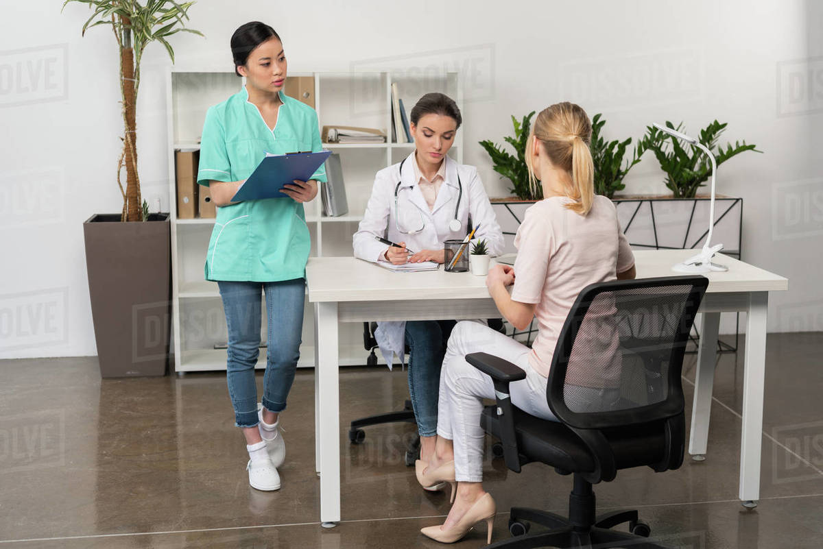 Blonde patient with doctor and internist sitting at table during ...