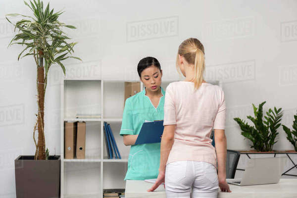 Back view of young patient standing near nurse writing in clipboard ...