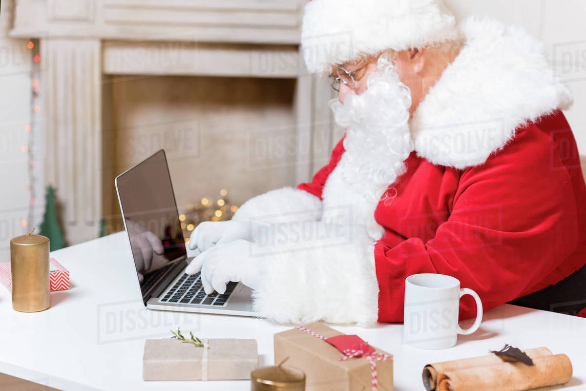 Side view of Santa Claus typing on laptop with blank screen at home ...