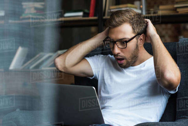 Young man sitting in armchair with surprised look on his face as he is ...