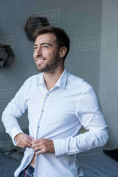 Half-length shot of young smiling man buttoning up a white shirt ...
