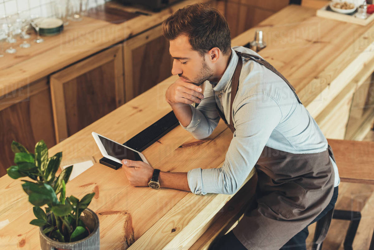 Side view of barista in apron using tablet while standing at counter in ...