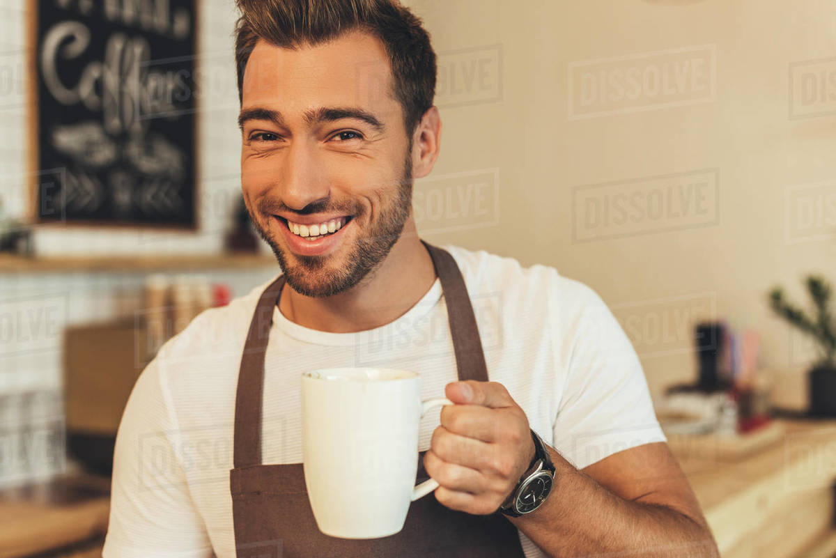 Portrait of cheerful barista with cup of coffee looking at camera ...