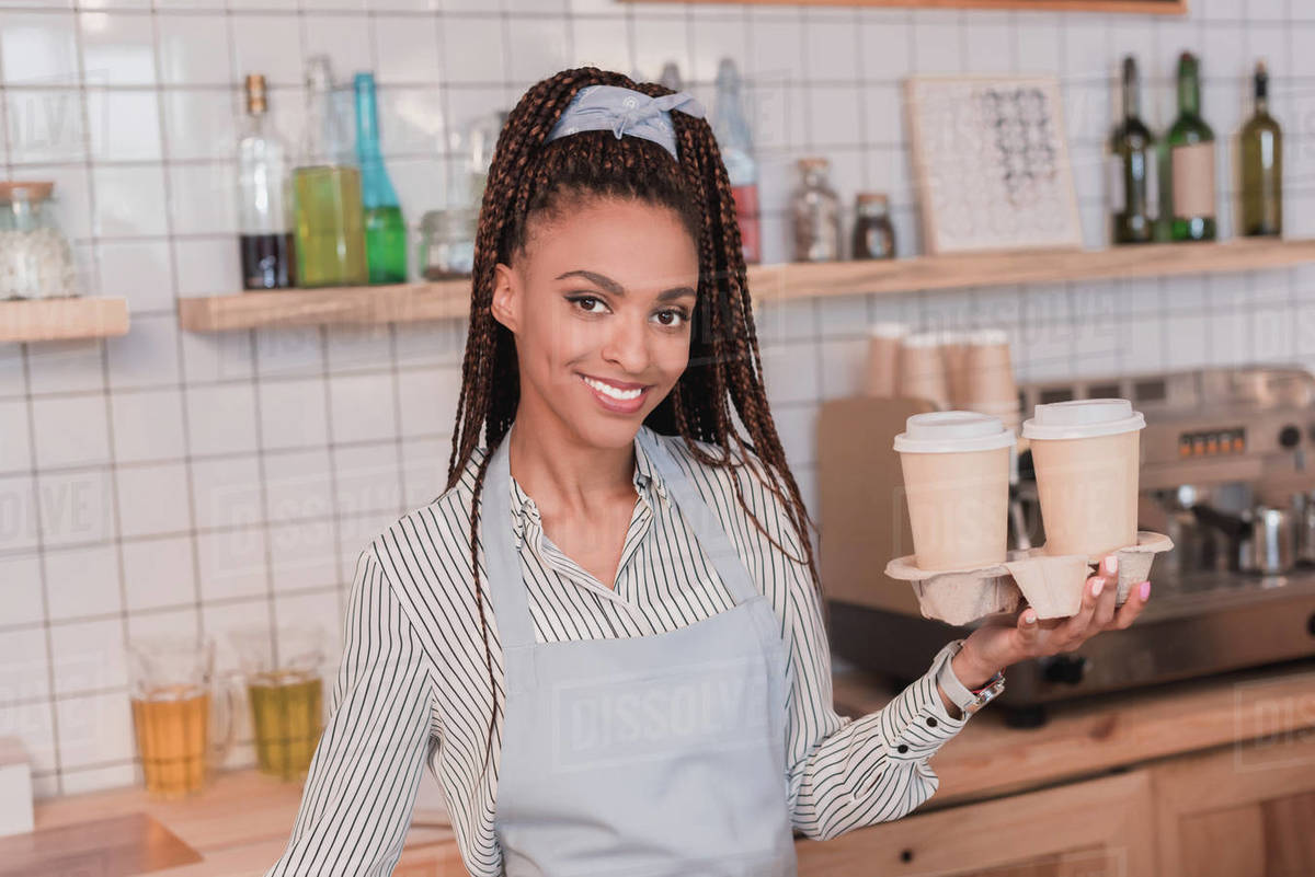 Smiling African American barista holding two paper cups in a holder at ...