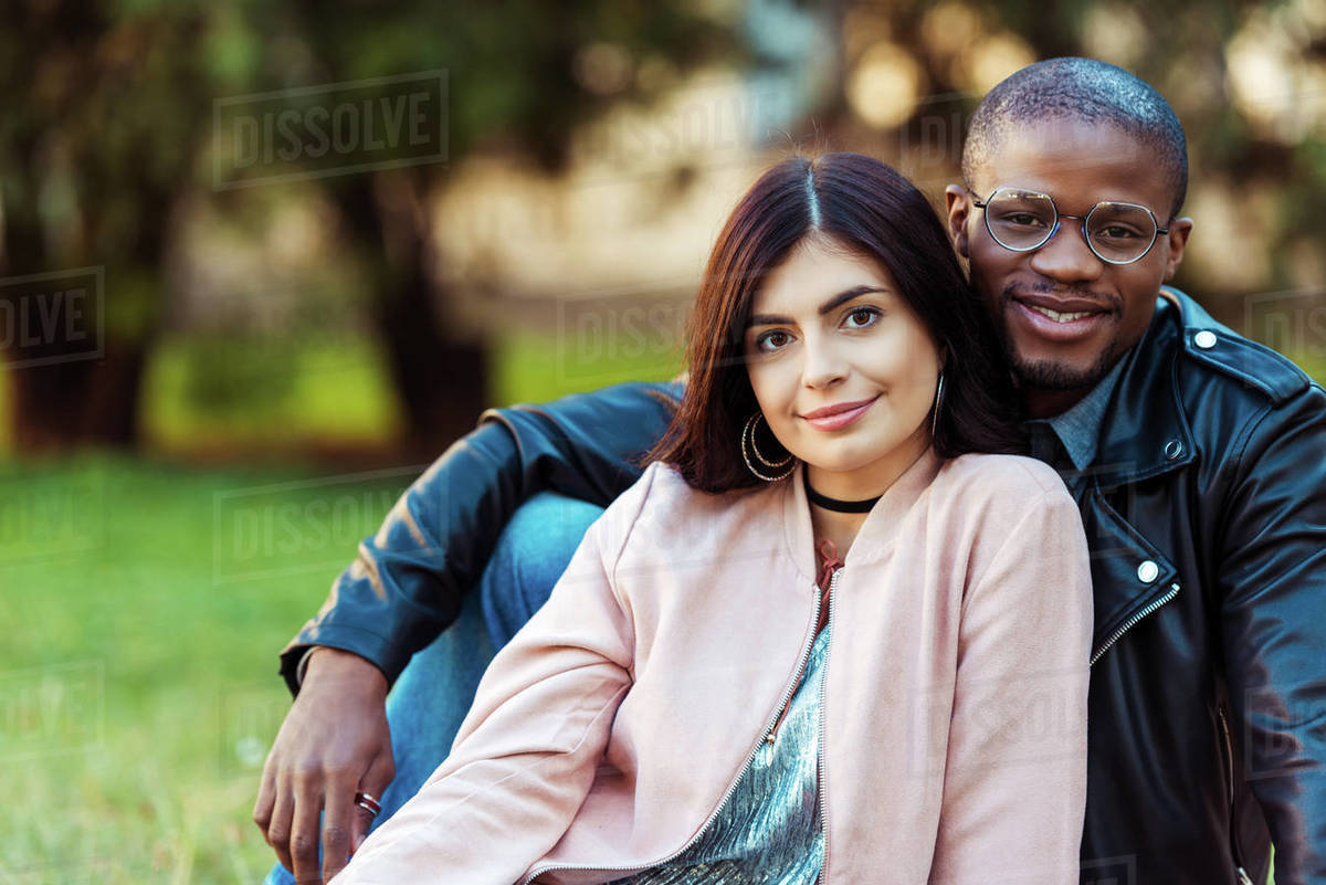 Portrait of lovely multicultural couple sitting on a grass in the park ...