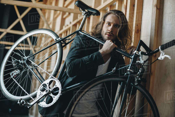 Stylish bearded young man with long hair carrying bicycle on shoulder ...
