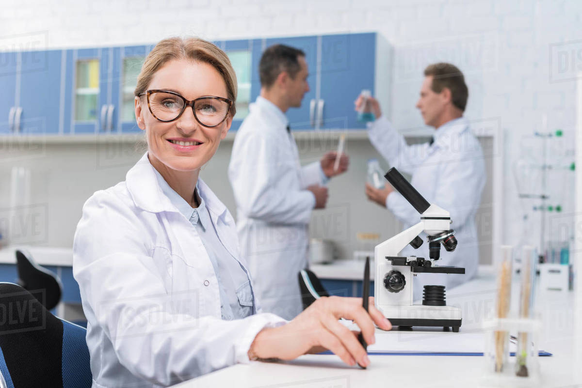 Beautiful scientist in eyeglasses sitting at table and smiling at ...