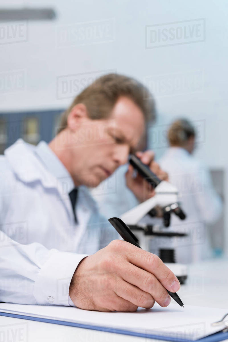 Concentrated lab technician taking notes while doing microscope sample ...