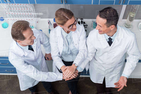 Team of smiling scientists in white coats, holding hands in laboratory ...