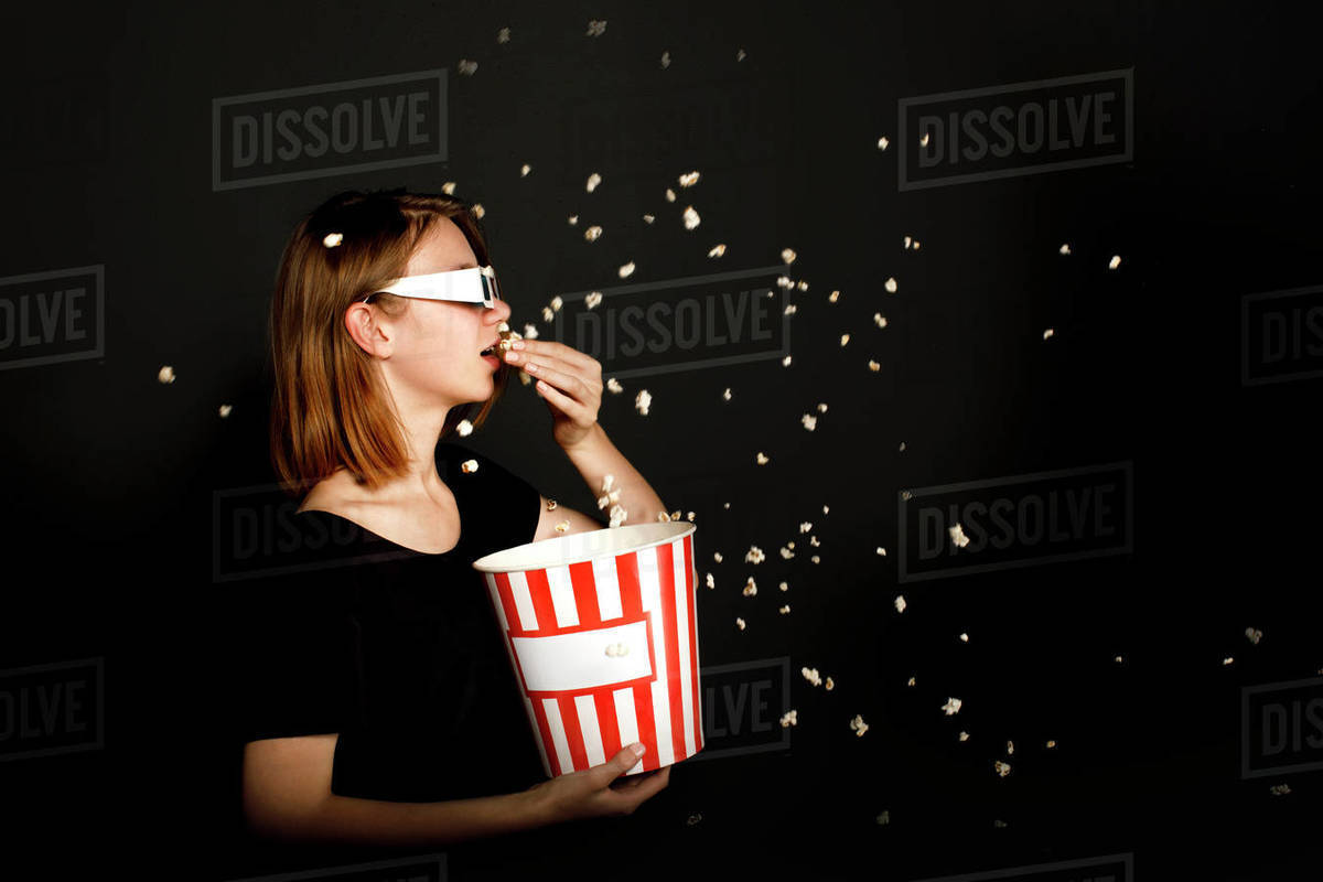 Side view of young woman in 3d glasses eating popcorn isolated on black ...