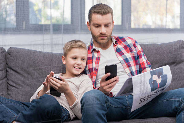 Father with newspaper and little son using smartphones together at home ...