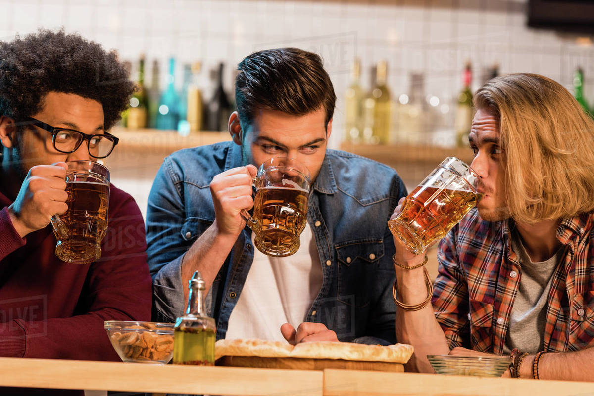 Three young multiethnic men drinking beer together in bar - Royalty ...
