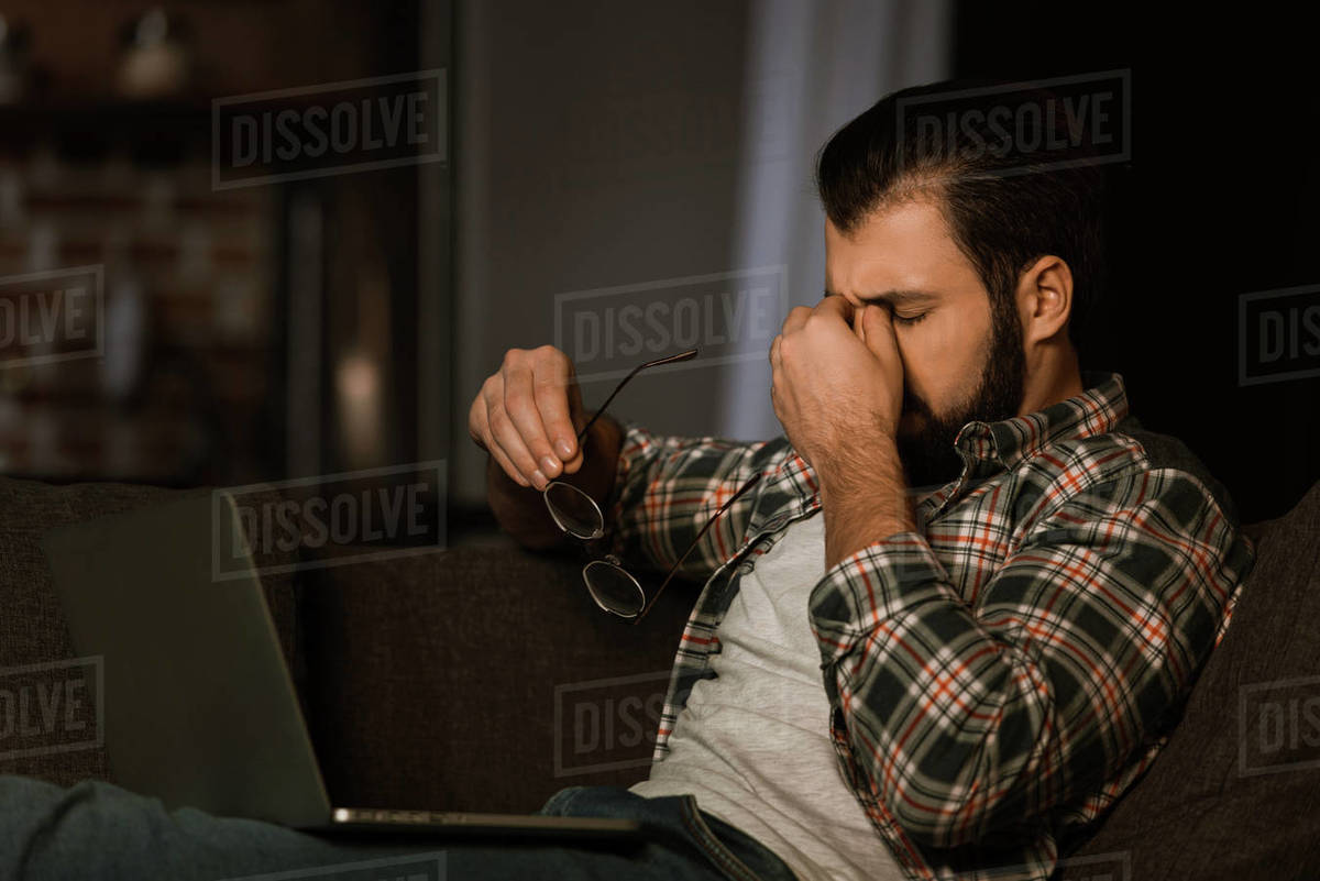 Tired man with glasses sitting on couch with laptop and rubbing head ...