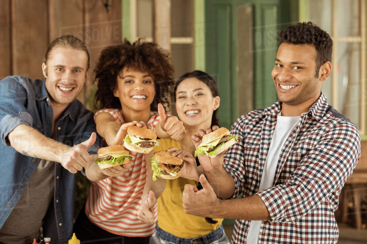 Young smiling multiethnic friends holding burgers at picnic on patio ...