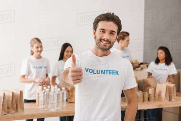 Young handsome volunteer showing thumb up with blurred colleagues ...